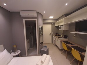Quarto de hotel compacto com uma cama, uma escrivaninha, uma cadeira, uma mesinha, micro-ondas, televisão e ar condicionado.