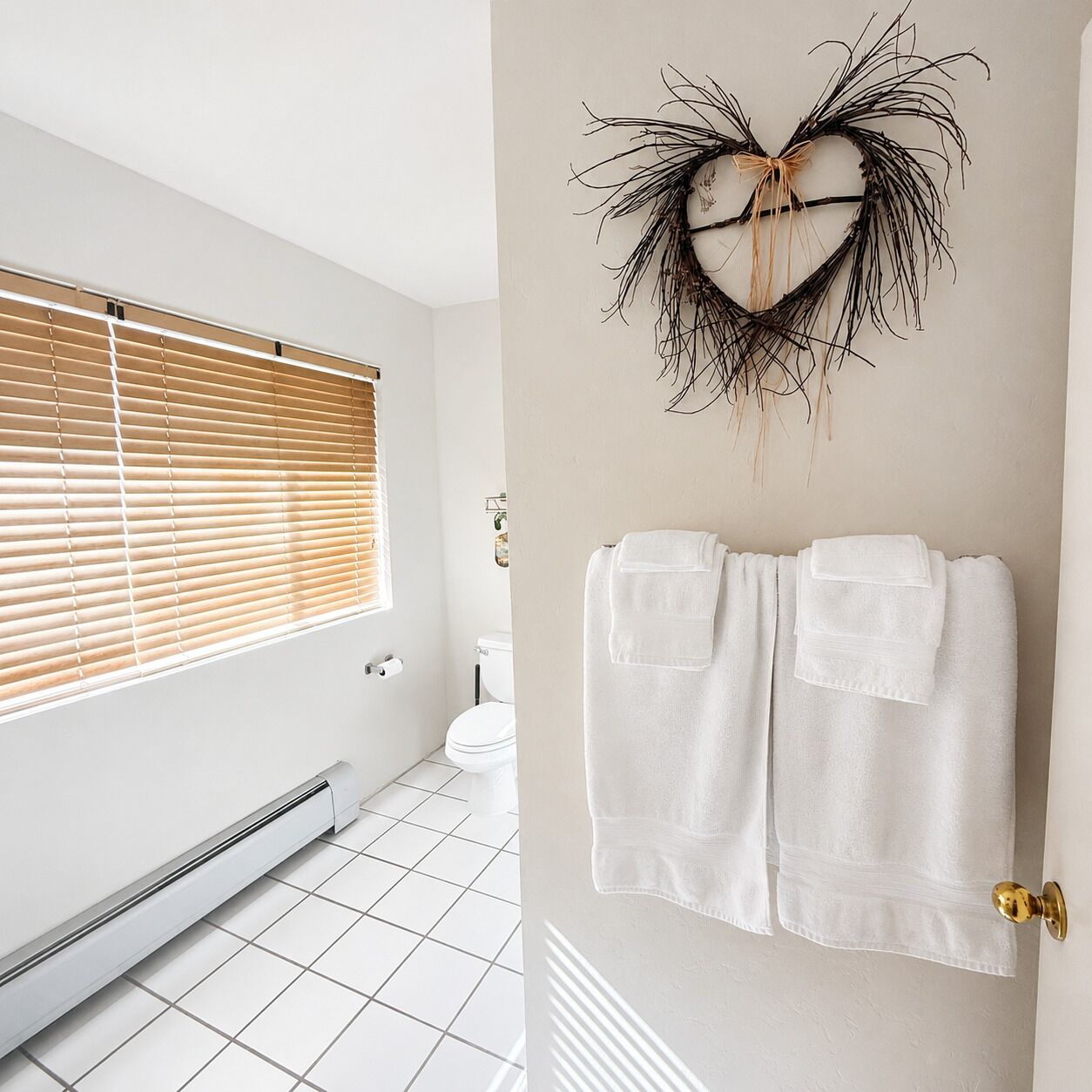 Um banheiro com piso de azulejos brancos, vaso sanitário, porta-toalhas fixado na parede com duas toalhas e uma coroa decorativa em formato de coração na parede.