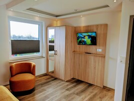 Quarto moderno com um armário de madeira, televisão fixada na parede, janela com vista para árvores e uma poltrona de couro marrom.