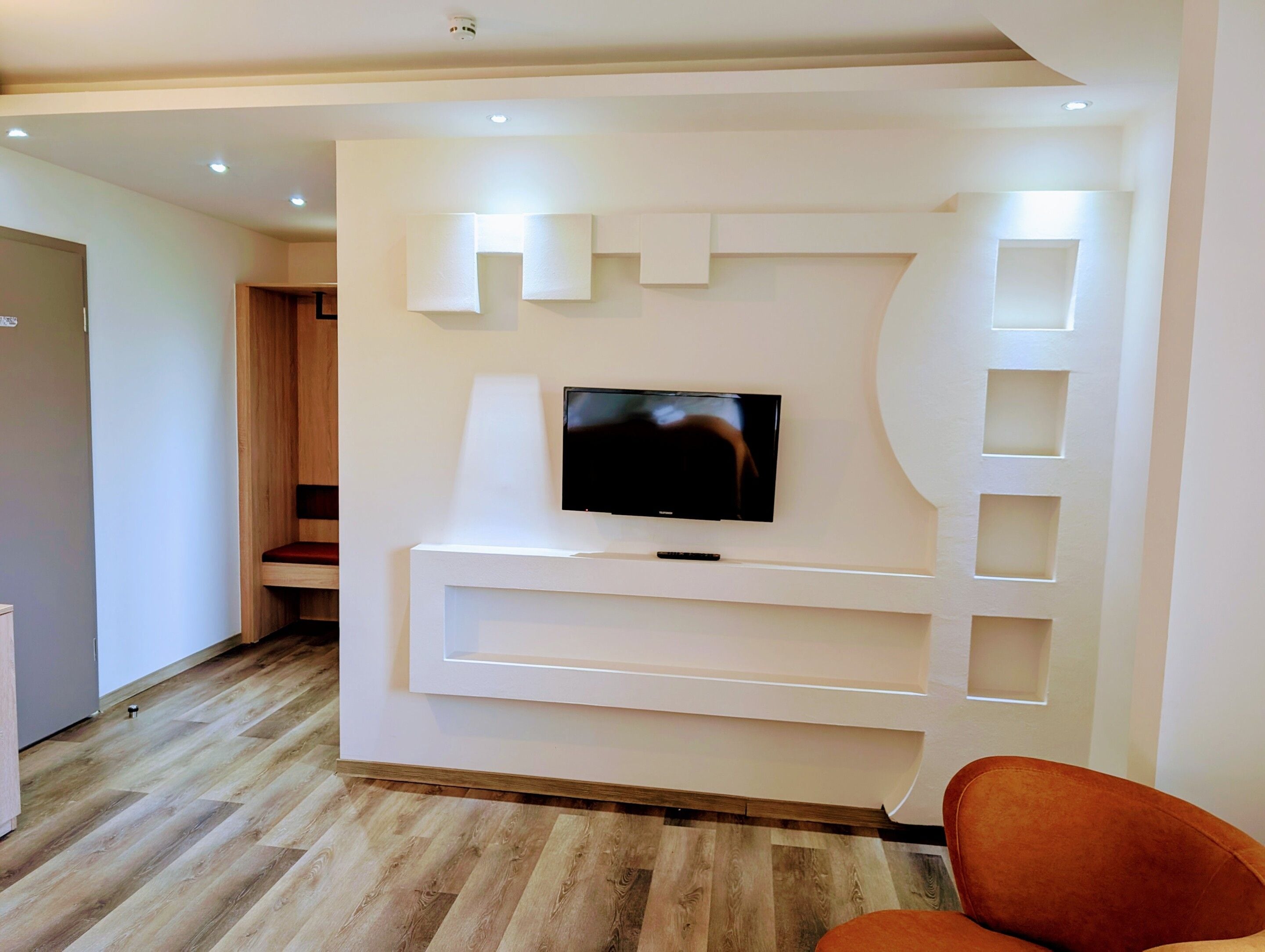 Uma sala de estar moderna com uma TV de tela plana presa a uma parede branca, piso de madeira e uma poltrona vermelha.