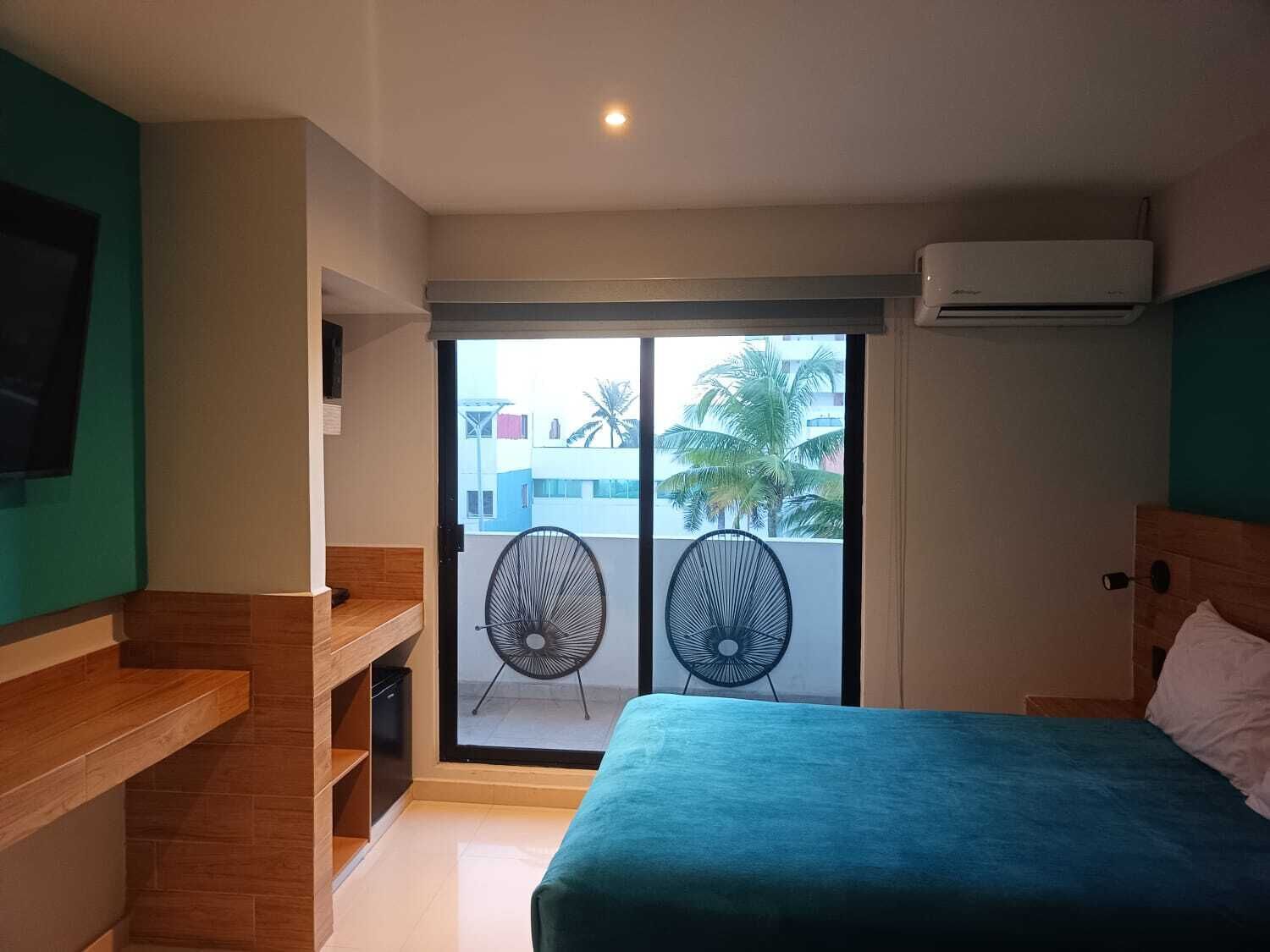 Quarto com cama, televisão, varanda com cadeiras e ar condicionado.