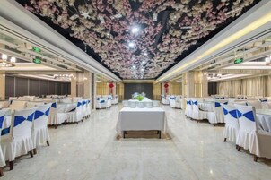 Salão de festas com mesas dispostas em fileiras, decoração floral no teto e arranjo floral central.