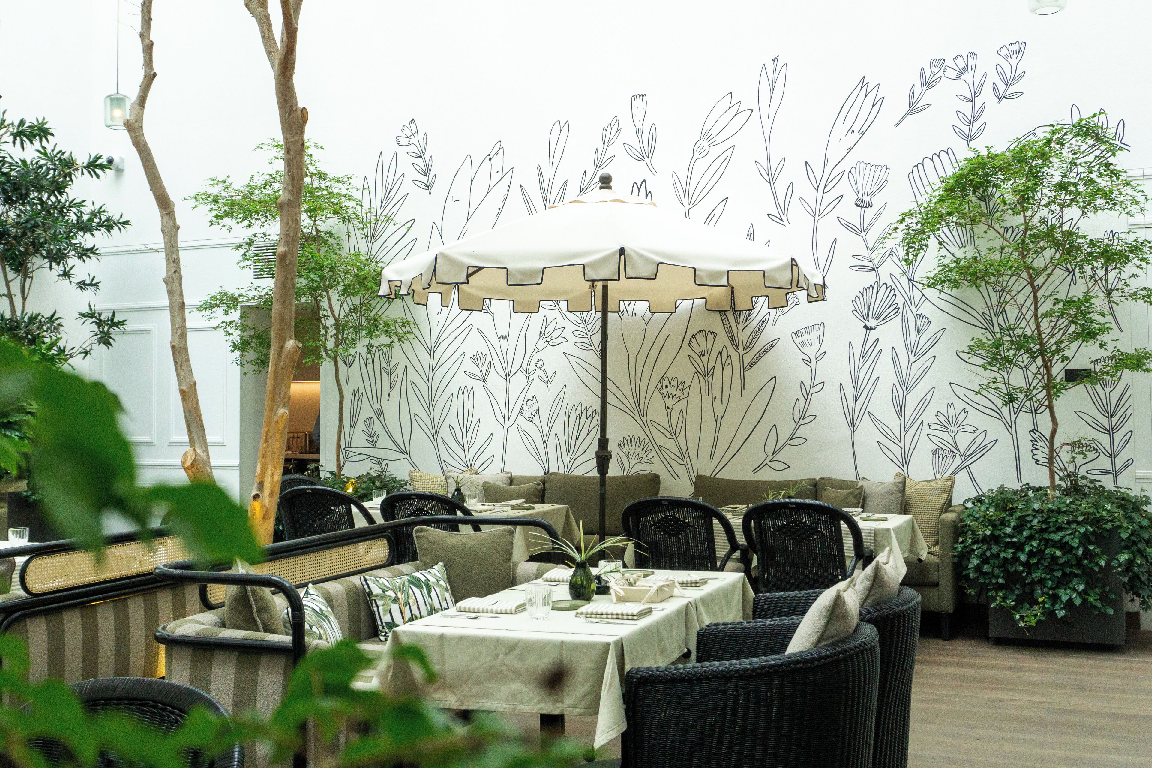 Uma área de refeições ao ar livre com móveis de vime, uma mesa com toalha branca e uma parede com mural floral.