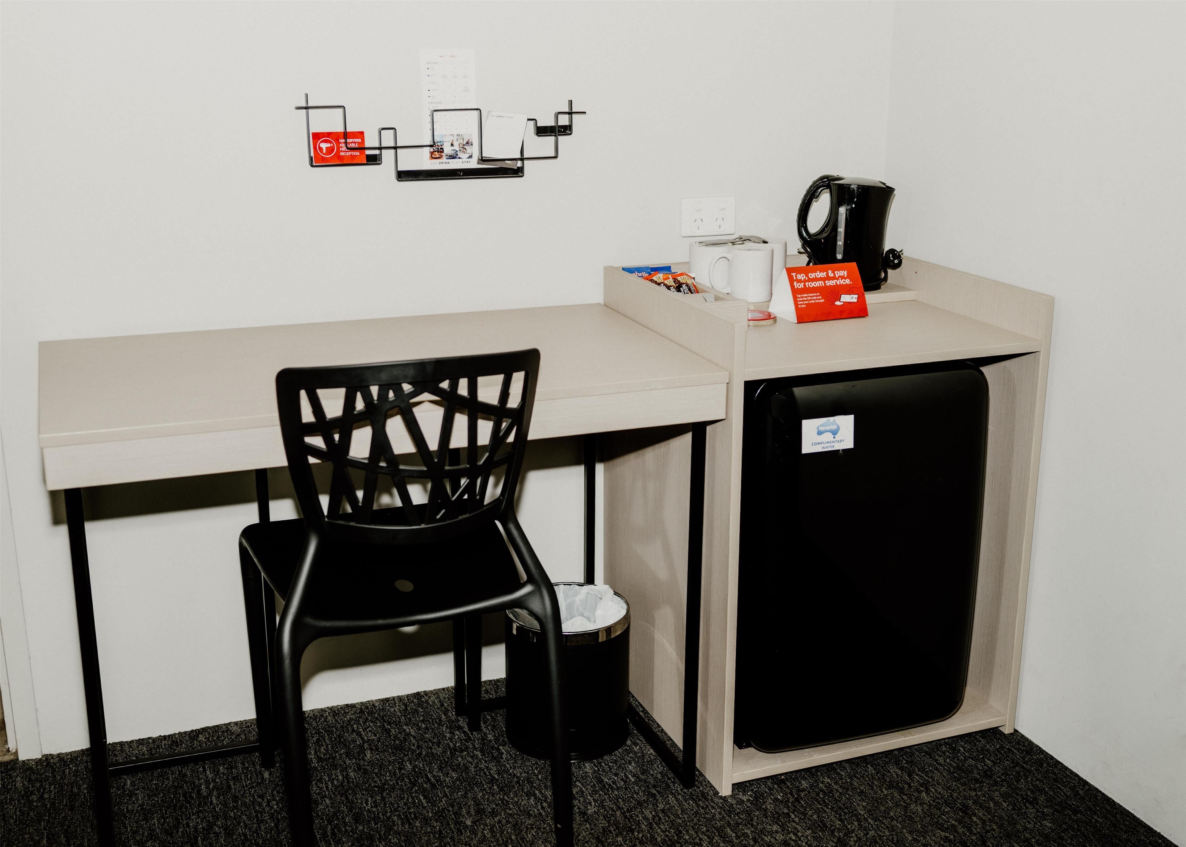 Quarto de hotel compacto com cozinha compacta, uma cadeira preta, um pequeno frigorífico e uma máquina de café.