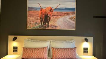 Quarto de hotel com uma cama grande, duas almofadas estampadas, um quadro de uma vaca das Terras Altas na parede e luminárias de cabeceira.