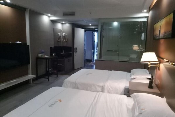 Quarto de hotel com duas camas, uma escrivaninha, televisão e banheiro com chuveiro de vidro.