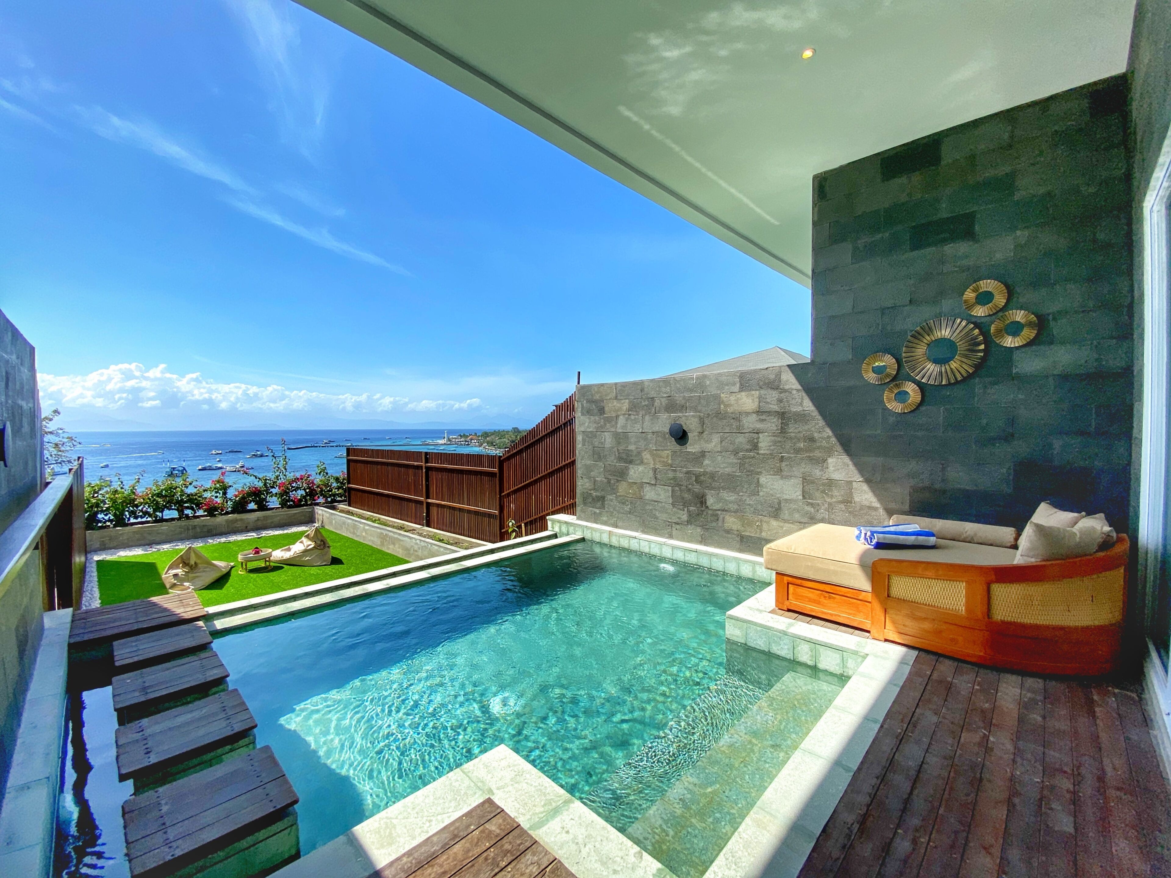 Uma piscina infinita moderna com vista clara para o oceano e barcos, um deck de madeira e uma área de estar aconchegante com almofadas.