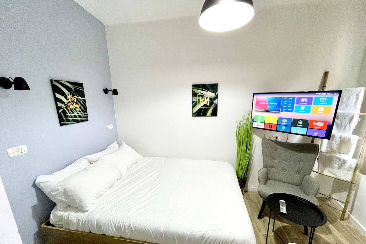 Quarto de hotel moderno com cama, televisão presa à parede, uma mesa pequena e uma cadeira.
