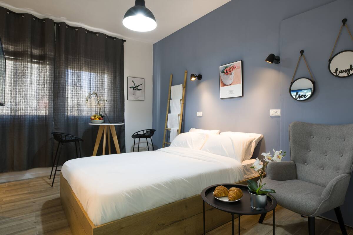 Quarto moderno com cama grande, poltrona cinza, mesa pequena com bandeja de croissants e um vaso de planta.