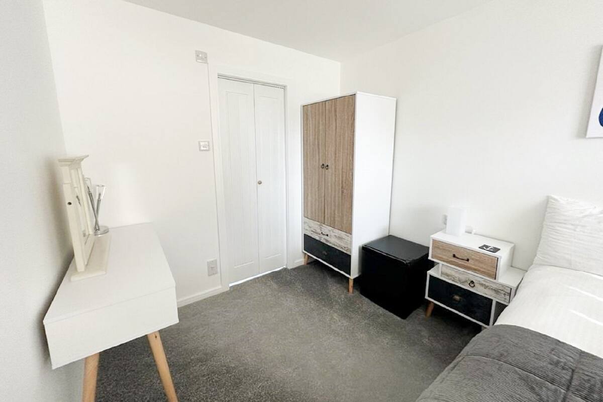 Quarto com cama, guarda-roupa, mesa de cabeceira e pequena escrivaninha.