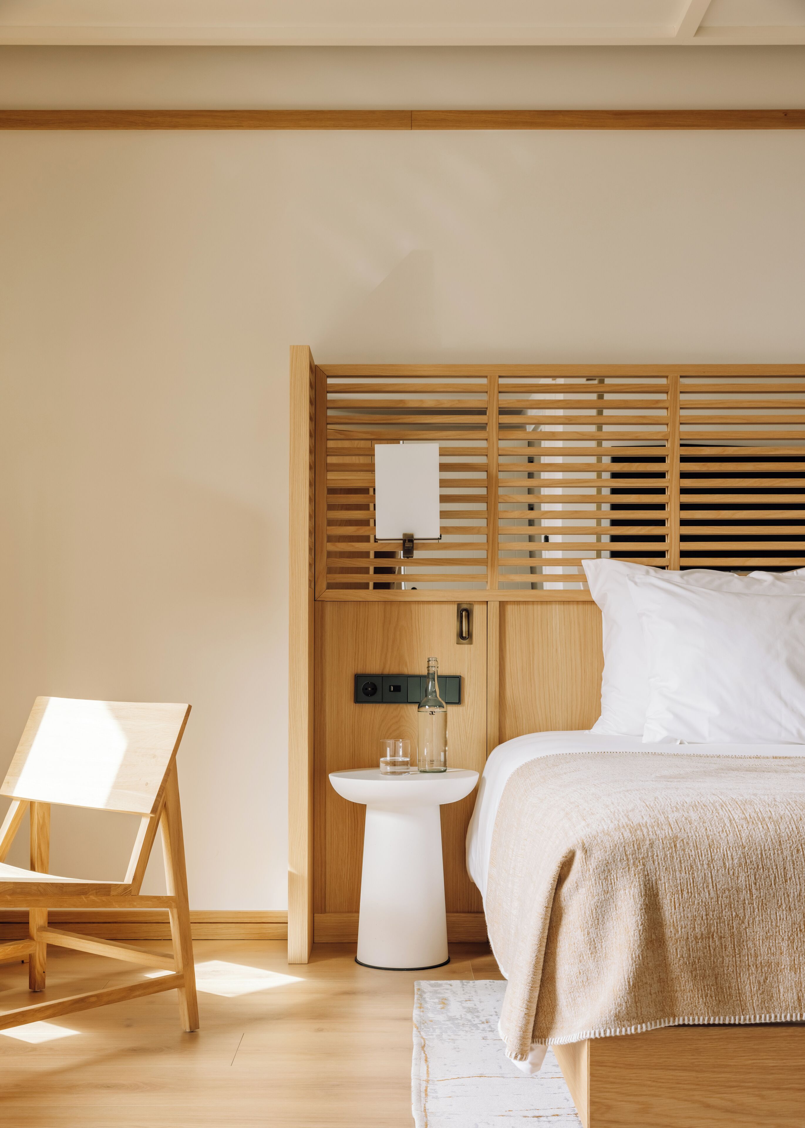 Quarto minimalista com cabeceira de madeira, mesa de cabeceira com garrafa e copo, pequena mesa redonda branca e uma cadeira de madeira.