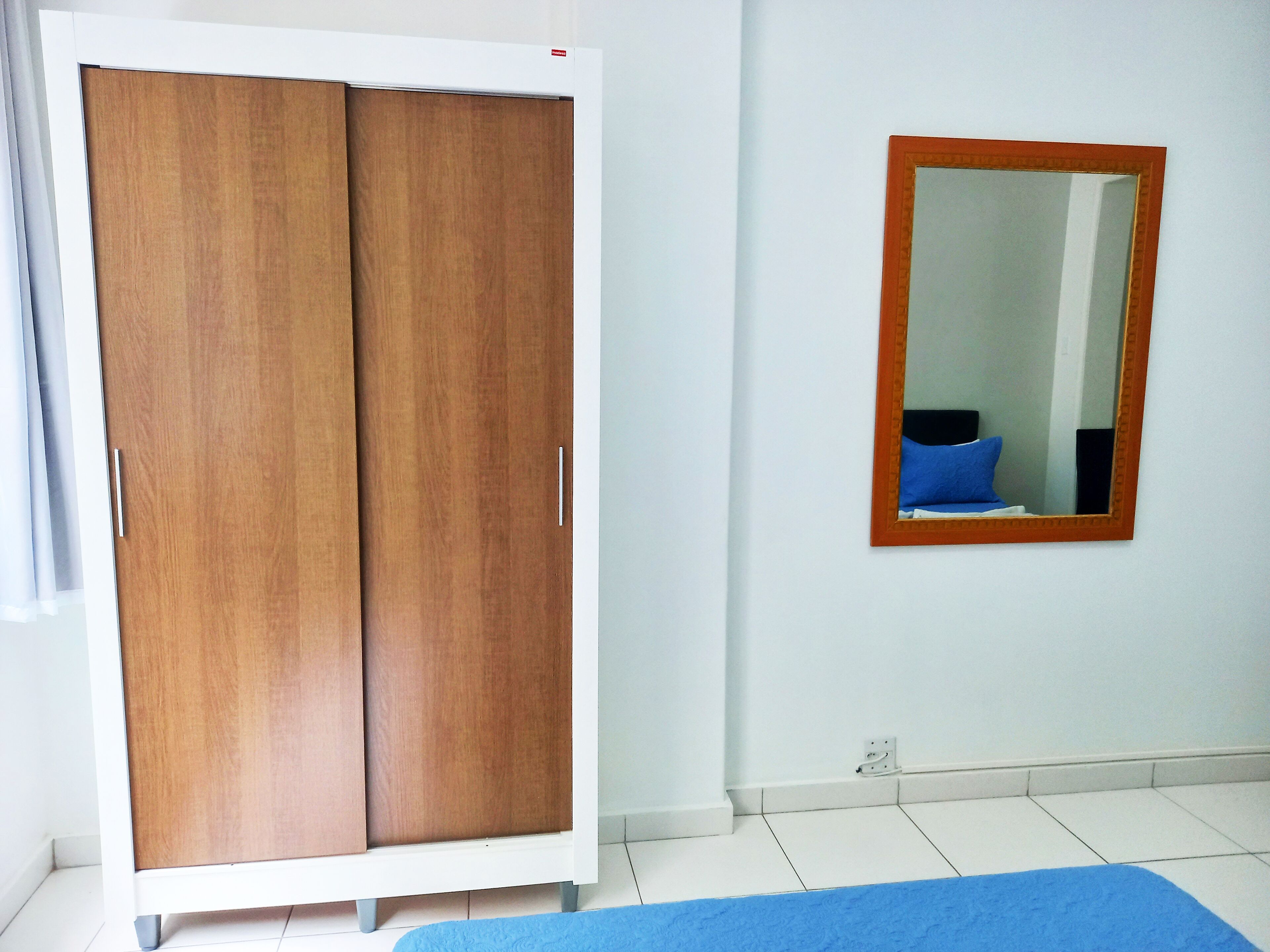 Quarto com guarda-roupa de madeira, espelho na parede e tapete azul no chão.