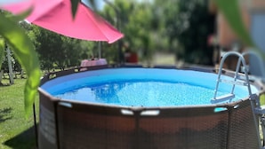 Piscina semissubterrânea com escada, rodeada por vegetação e um guarda-sol rosa.