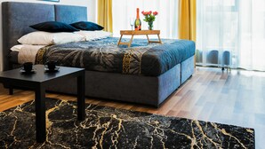 Quarto moderno com uma cama grande, uma mesinha de cabeceira, uma pequena mesa com bebidas e vista da cidade através de amplas janelas.