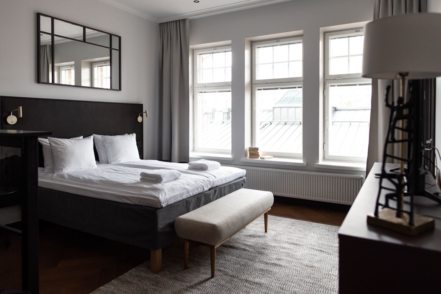 Quarto de hotel moderno com uma cama grande, mesa de cabeceira, um banco e vista da cidade através de amplas janelas.