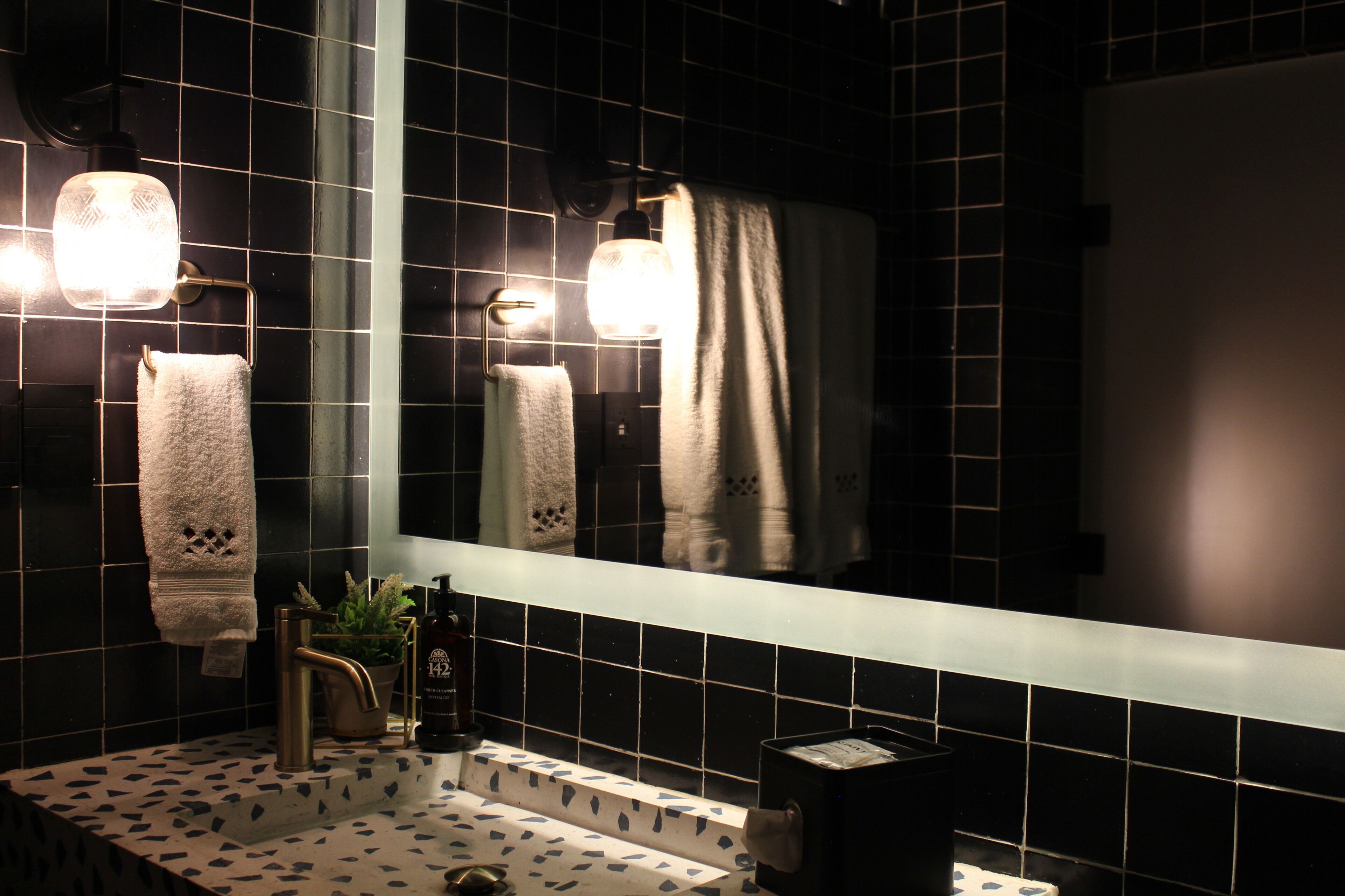 Casa de banho com paredes revestidas de azulejos pretos, uma pia branca com torneira dourada e três apliques de parede iluminados.