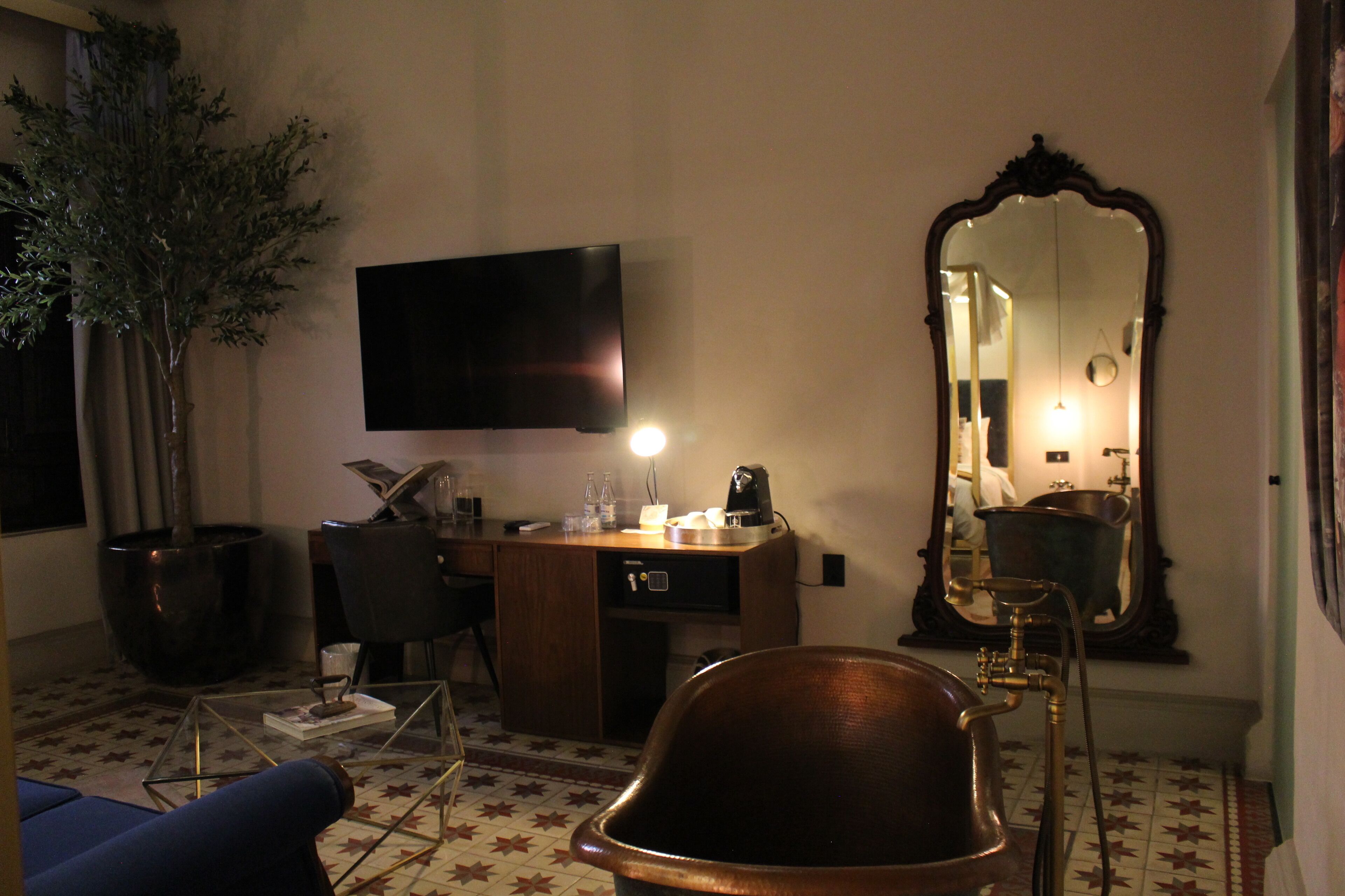 Quarto com televisão de tela plana na parede, escrivaninha com computador e máquina de café, espelho grande e planta em vaso.