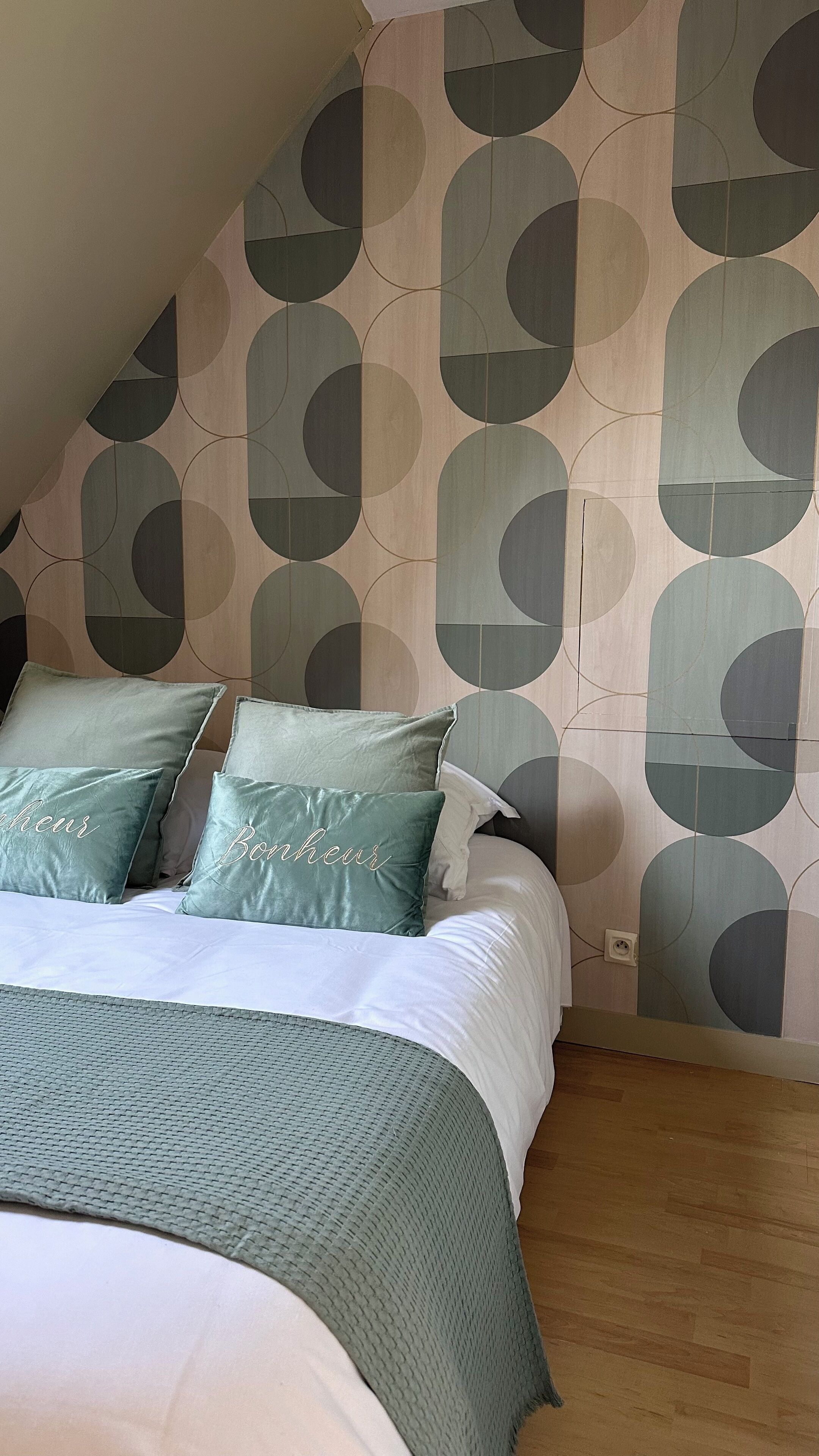Um quarto com teto inclinado, uma cama com colcha branca e verde, duas almofadas com texto, e uma parede com padrão geométrico.
