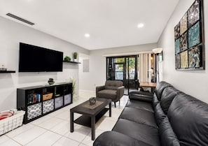 Uma sala de estar moderna com televisão de tela plana, sofá de couro preto, poltrona cinza, mesa de centro e estante de livros.