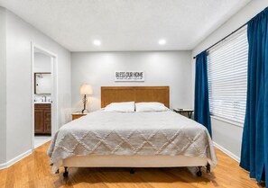 Quarto com cama, mesa de cabeceira, luminária, placa na parede e janela com cortinas.