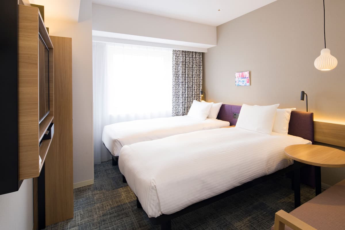 Quarto de hotel com duas camas, uma mesinha, uma cadeira, uma escrivaninha e uma janela com cortinas.