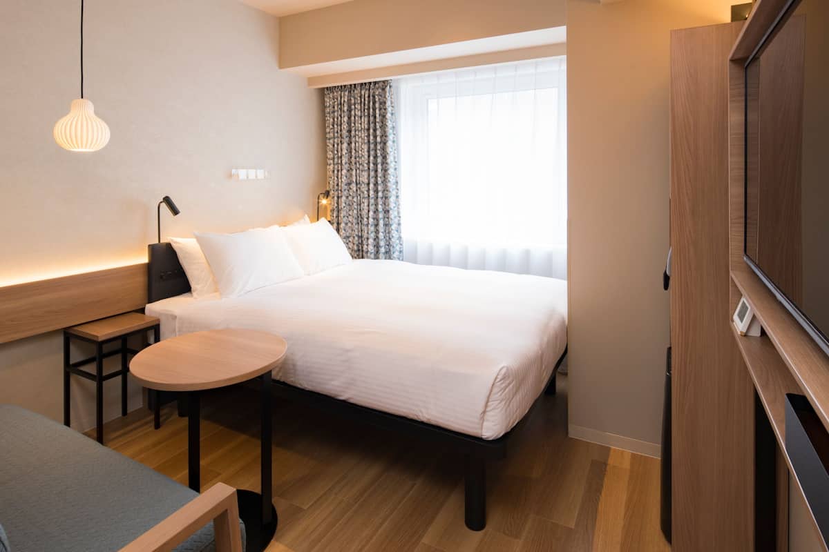 Quarto de hotel com cama, mesa de cabeceira, abajur, televisão e vista para o exterior pela janela.