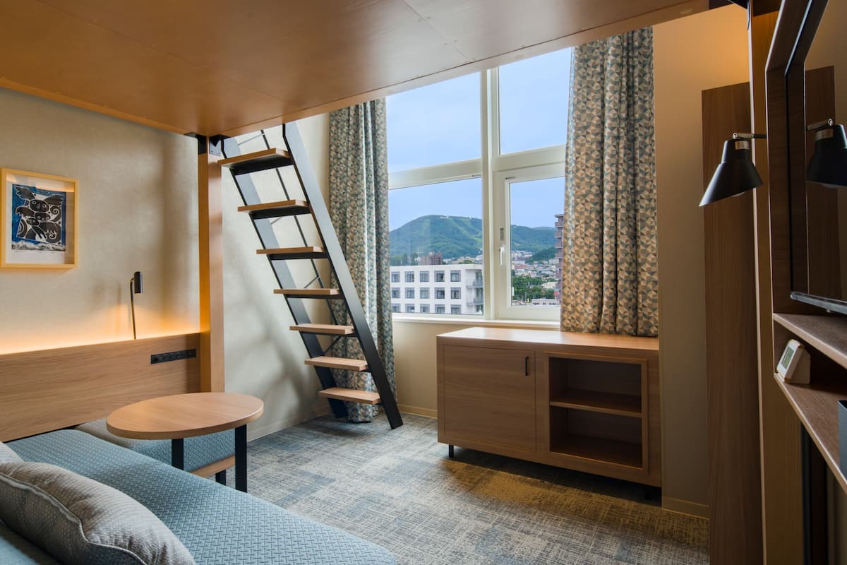 Quarto de hotel compacto com sofá, uma pequena mesa, escrivaninha com cadeira e escada que leva a uma janela, com vista para edifícios e colinas.