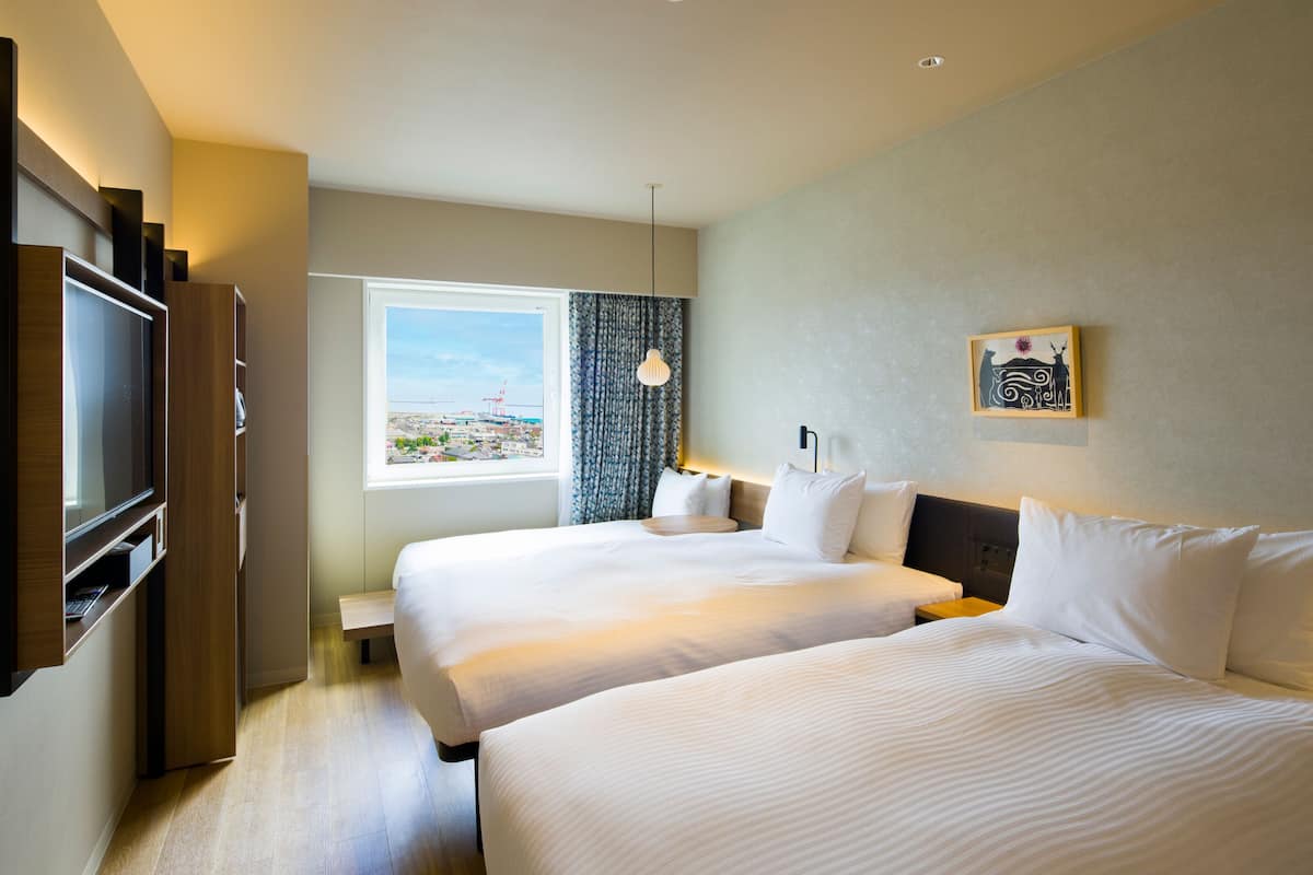 Quarto de hotel com duas camas, uma televisão, uma janela com vista para a cidade e um quadro na parede.