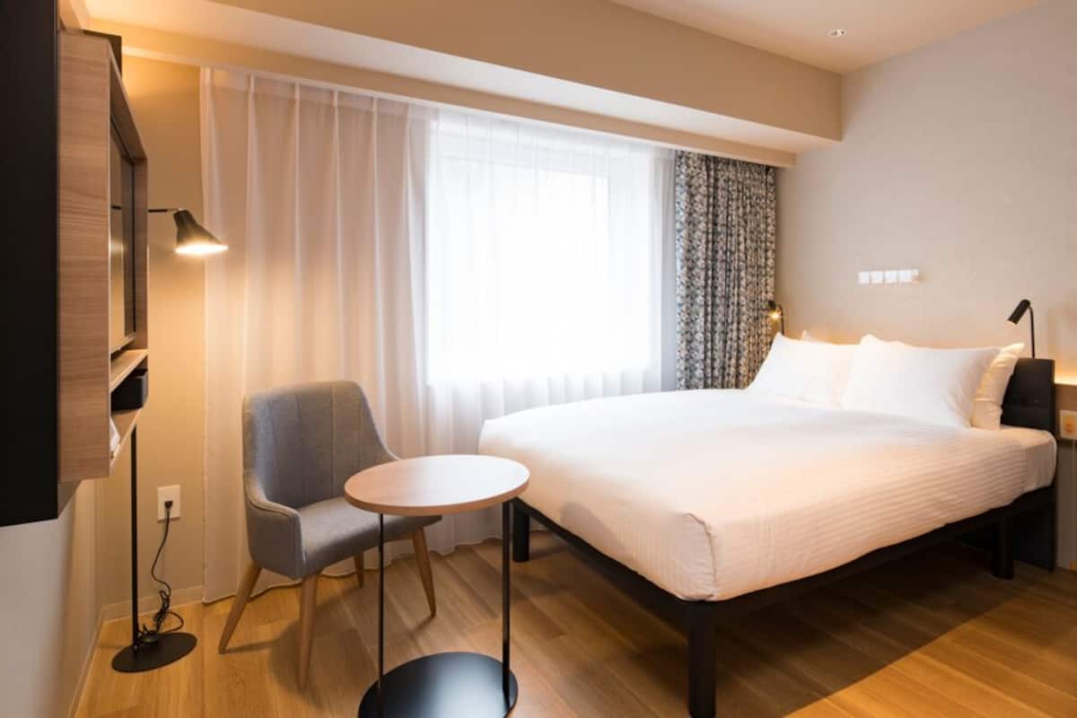 Quarto de hotel com cama, uma cadeira, mesa pequena, escrivaninha e televisão.