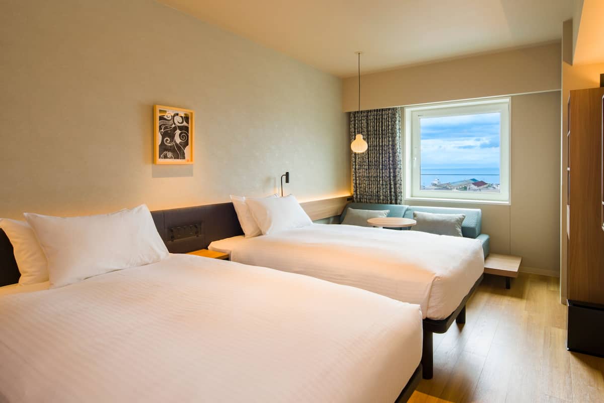 Quarto de hotel com duas camas, uma mesinha, vista para o mar e um quadro na parede.