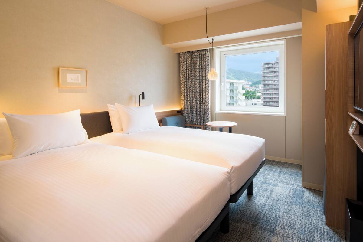 Quarto de hotel com uma cama grande, uma mesinha, uma janela com vista para edifícios e um guarda-roupa.