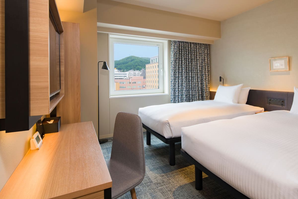 Quarto de hotel com duas camas, uma escrivaninha, uma cadeira, uma janela com vista para edifícios e uma cortina.