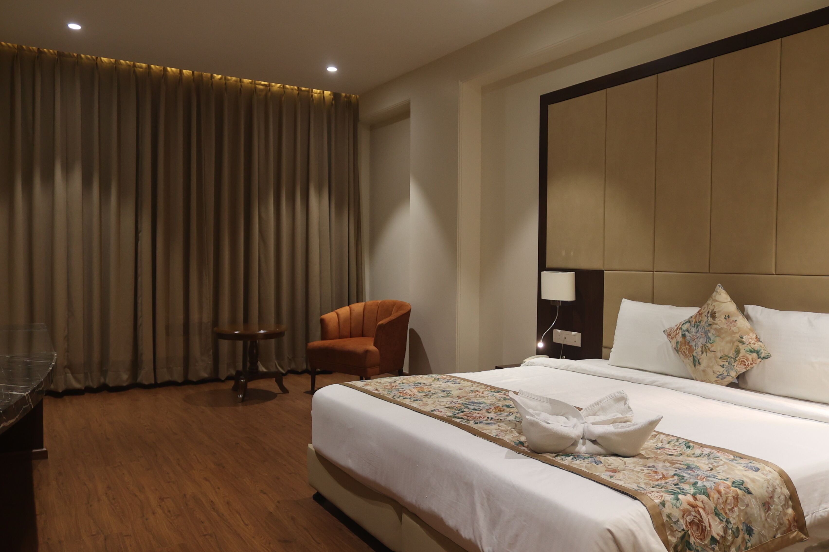 Quarto de hotel com uma cama grande, piso de madeira, uma cadeira e uma cortina.