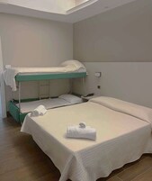 Quarto com beliche de estrutura verde e roupa de cama branca. Há uma pequena mesa de cabeceira com um abajur e uma toalha dobrada na cama.
