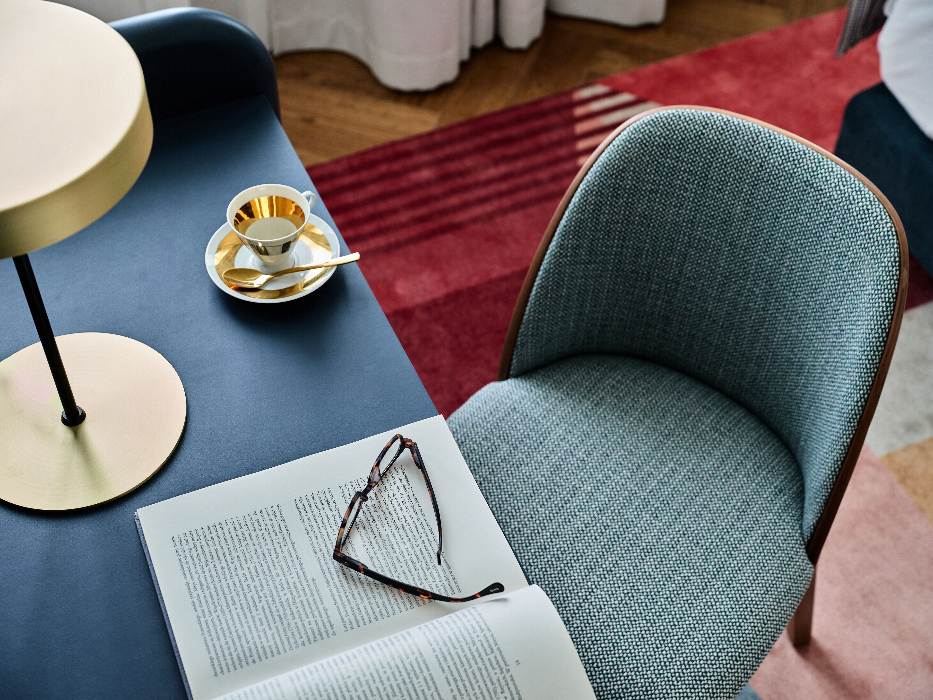 Quarto com uma poltrona azul, uma mesa com um livro e óculos, e uma xícara de café em um pires.