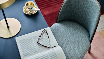 Quarto com uma poltrona azul, uma mesa com um livro e óculos, e uma xícara de café em um pires.