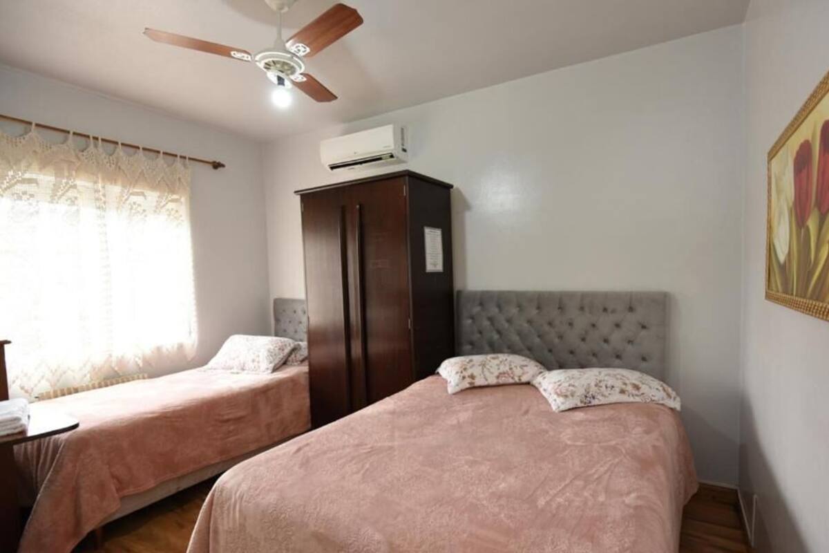 Quarto com duas camas, um guarda-roupa, ar condicionado, ventoinha de teto e uma janela com cortinas de renda.