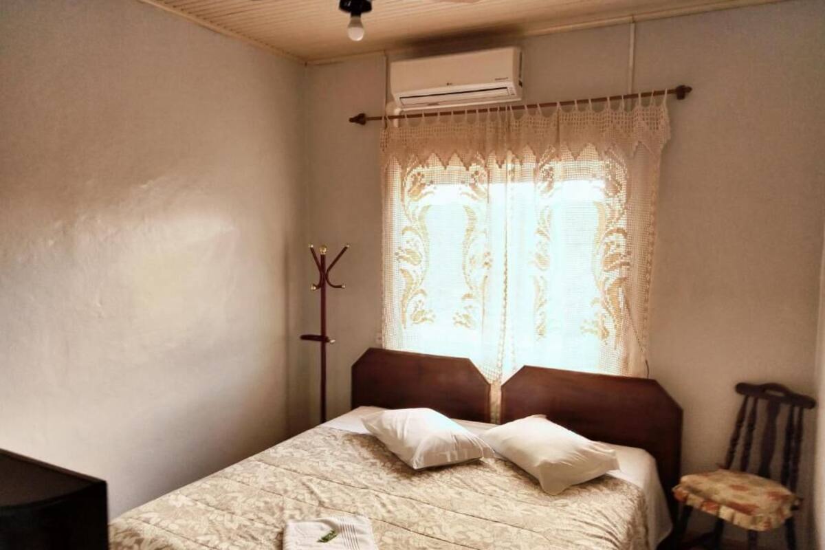 Quarto com cama, cadeira, janela com cortinas de renda e aparelho de ar condicionado.
