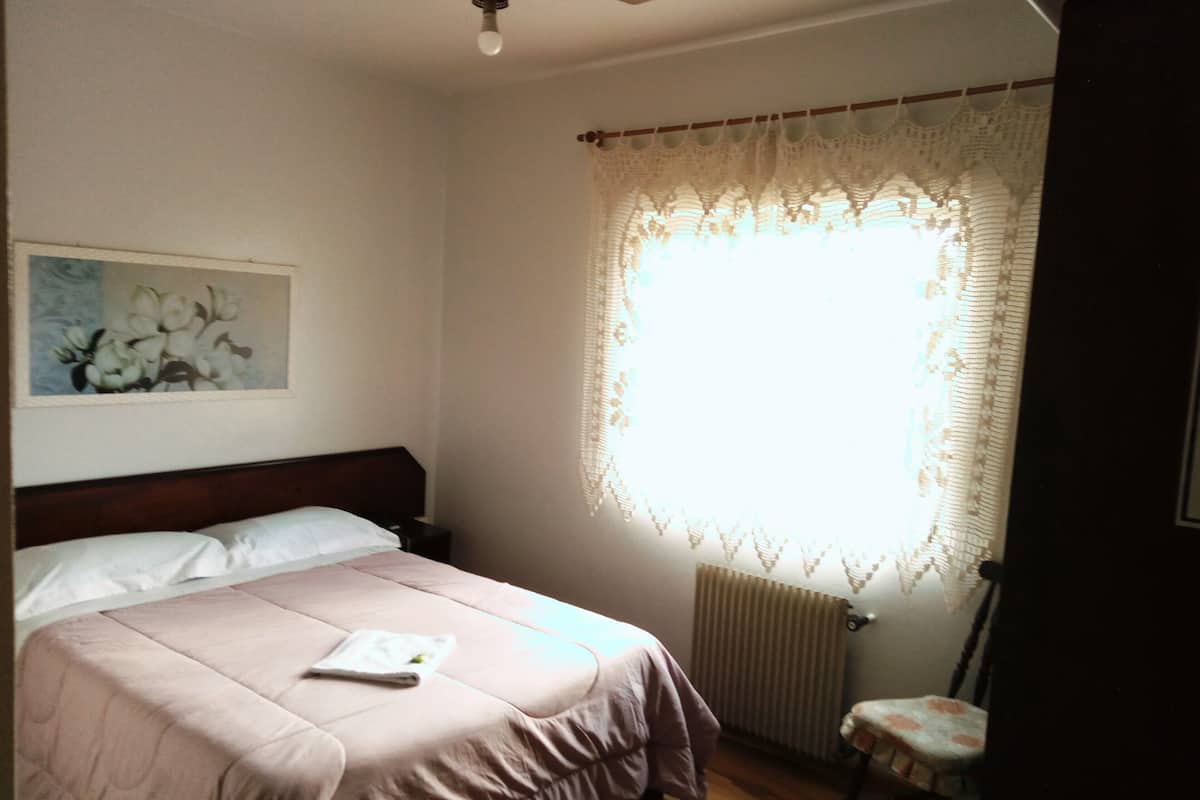 Quarto com cama, cadeira, radiador, ventoinha no teto e janela com cortinas de renda.