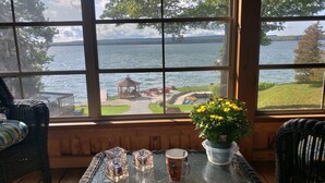 Quarto com uma grande janela com vista para um lago, móveis de vime, uma mesa de vidro com objetos decorativos e uma planta em vaso.