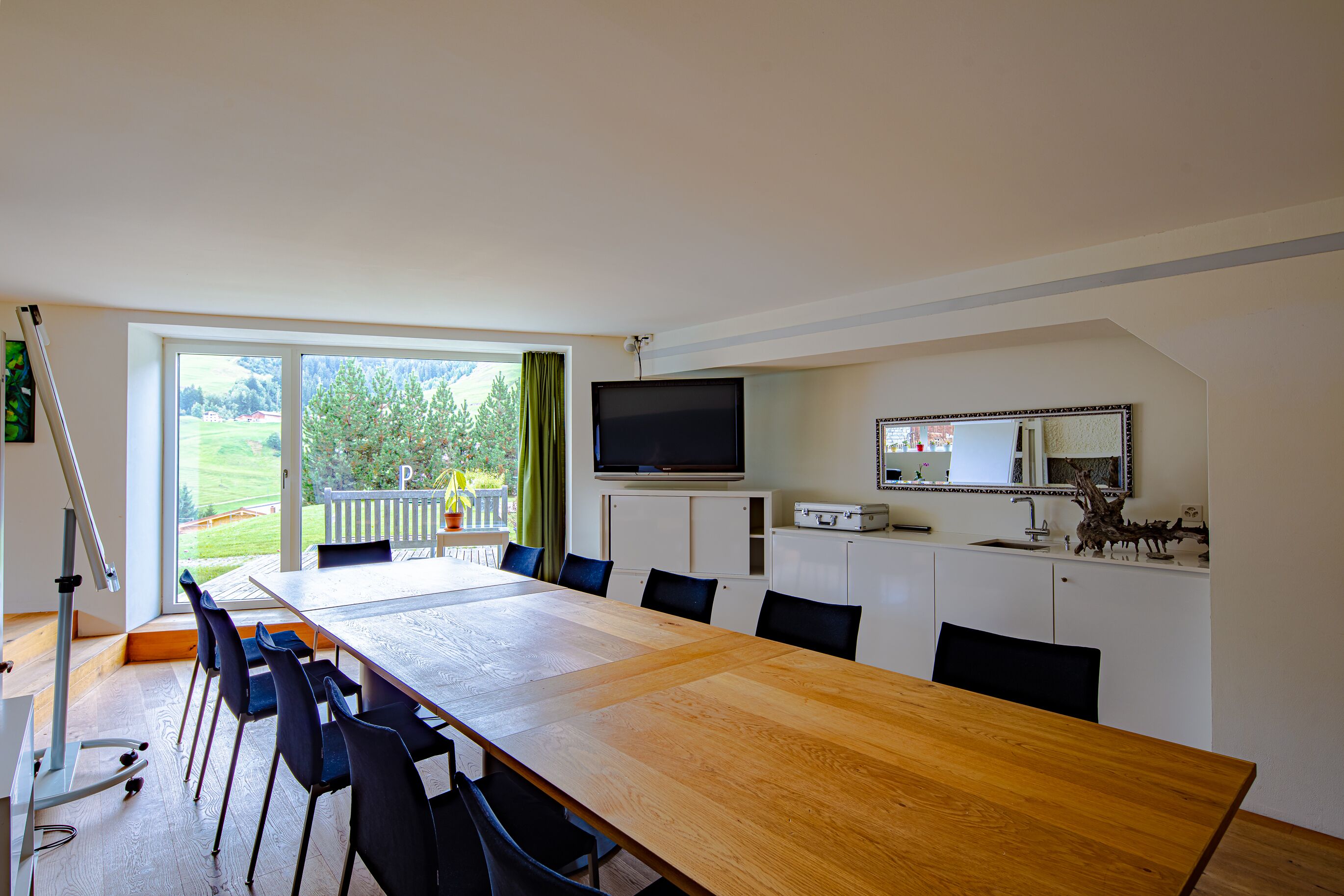 Uma sala de jantar espaçosa com uma longa mesa de madeira, cadeiras pretas, uma televisão montada acima e vista para um jardim através de grandes janelas.