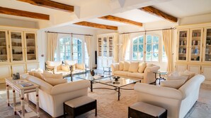 Uma sala de estar ampla, com tetos altos, vigas de madeira aparentes e grandes janelas. Possui vários espaços de estar com sofás e poltronas brancos, uma mesa de centro de vidro e estantes embutidas nas paredes.
