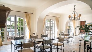Uma sala de jantar ampla com uma mesa grande e cadeiras, janelas grandes com cortinas e um candelabro.