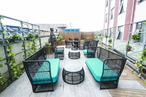 Uma área externa com assentos de vime e almofadas, cercada por plantas e um deck de madeira.