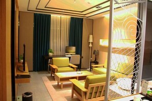 Quarto de hotel com escrivaninha, televisão, cama com cabeceira branca e área de estar com cadeiras e mesa.