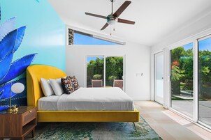 Quarto moderno com cama amarela, roupa de cama branca e uma grande janela com vista para o verde.