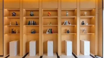 Um hall de hotel com design moderno, destacando um pilar central, diversas prateleiras com objetos decorativos e piso de mármore.