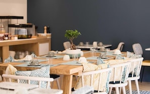 Uma área de refeições com uma mesa de madeira posta para uma refeição, cercada por cadeiras brancas com almofadas estampadas. Uma pequena planta em vaso está colocada sobre a mesa, e o ambiente possui um design moderno e minimalista, com uma parede escura ao fundo.