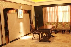 Quarto de hotel com mesa de jantar, quatro cadeiras, sofá, luminária e janela com cortinas.