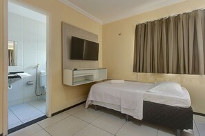 Quarto de hotel com cama, televisão na parede, banheiro visível por uma porta aberta e cortina na janela.